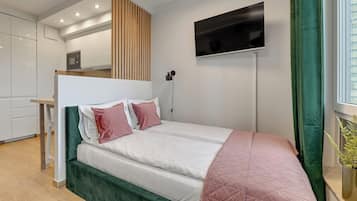 Quarto moderno com cama, TV fixada na parede e área de cozinha visível através de uma porta aberta.