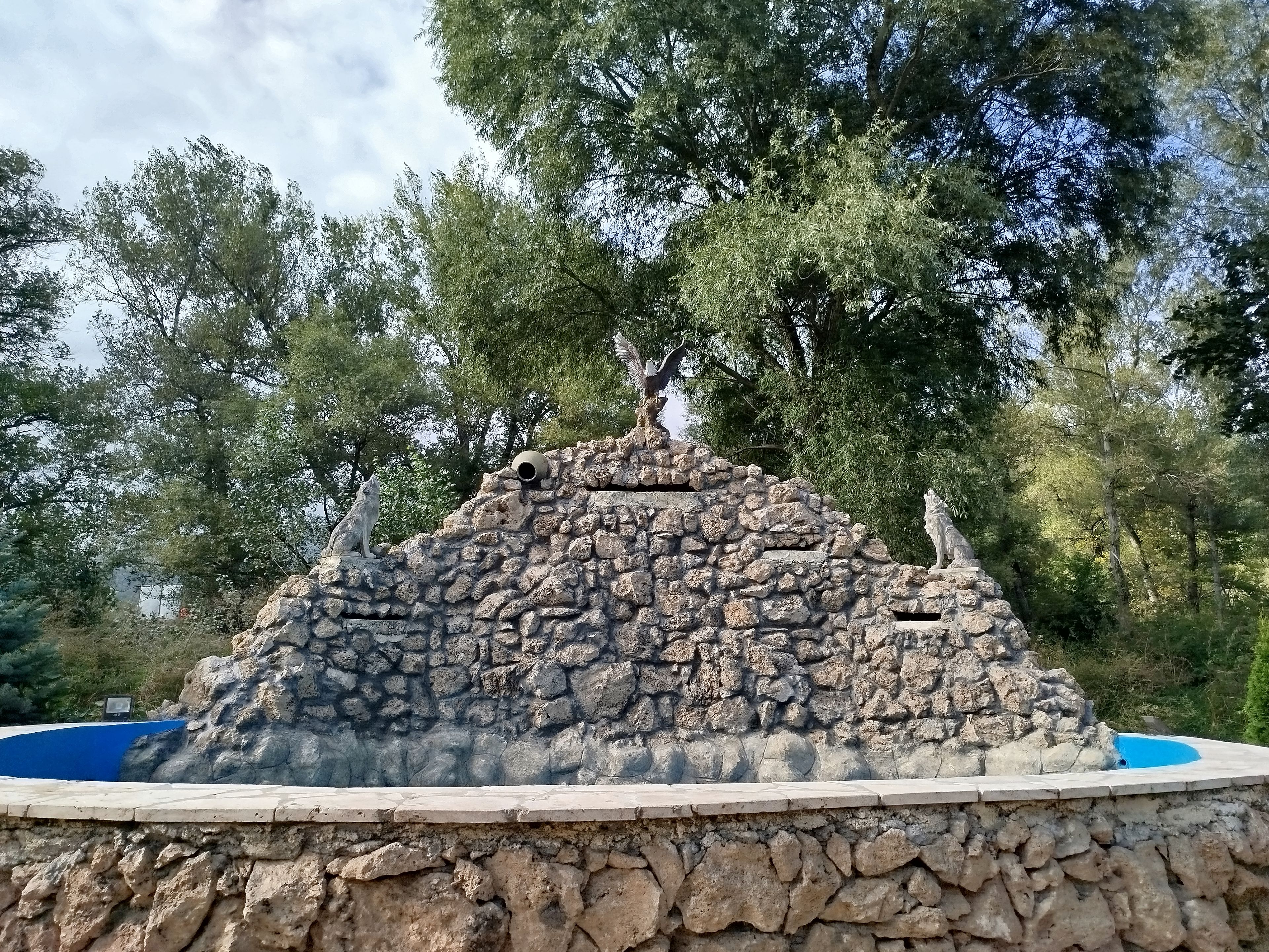 Uma fonte de pedra com duas estátuas de lobo, rodeada por árvores.