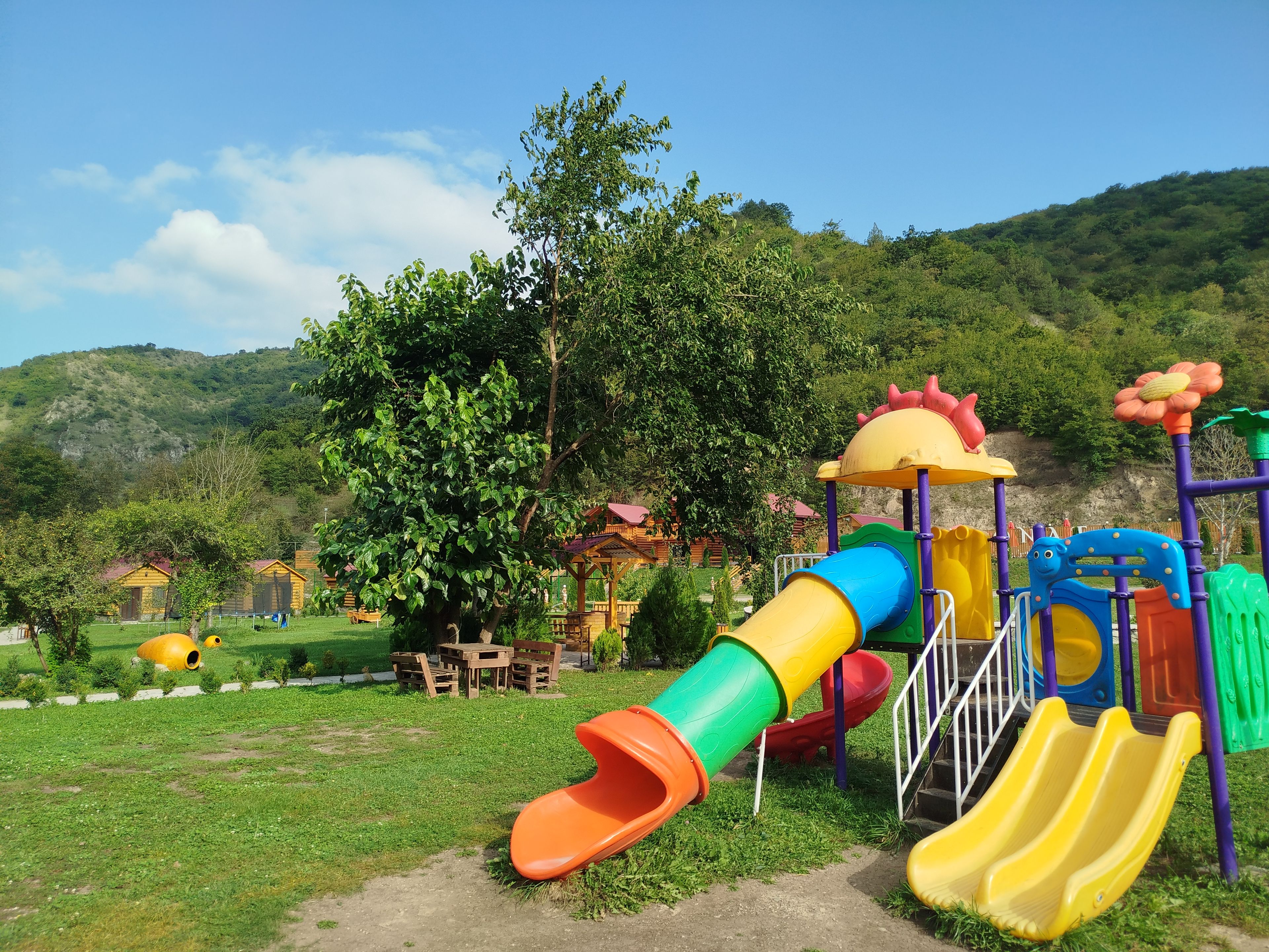 Parquinho com escorregadores e estruturas de escalada, cercado por vegetação e colinas.