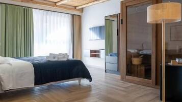 Quarto de hotel com piso de madeira, cama com colcha branca, cortina verde e uma janela grande.