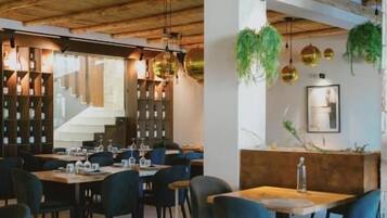 Interior de restaurante com vigas de madeira, mesas com cadeiras azuis e plantas penduradas.