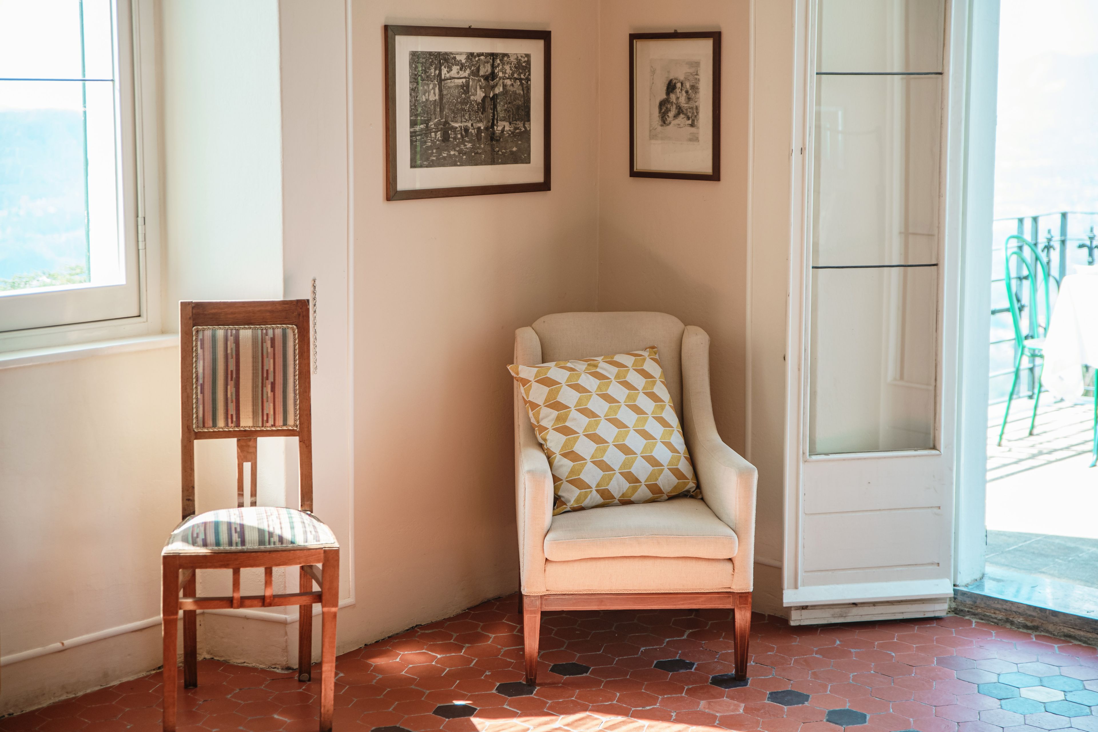 Um quarto com uma cadeira de madeira e uma poltrona estofada, piso ladrilhado e duas imagens emolduradas na parede.