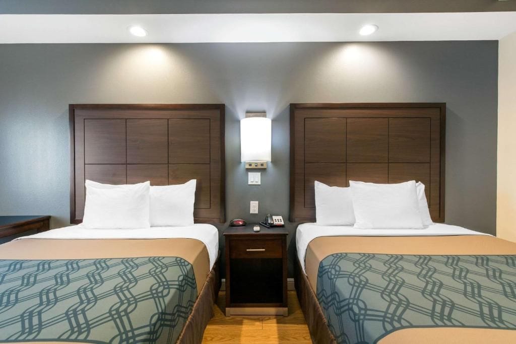 Quarto de hotel com duas camas, mesa de cabeceira e luminária fixa na parede.
