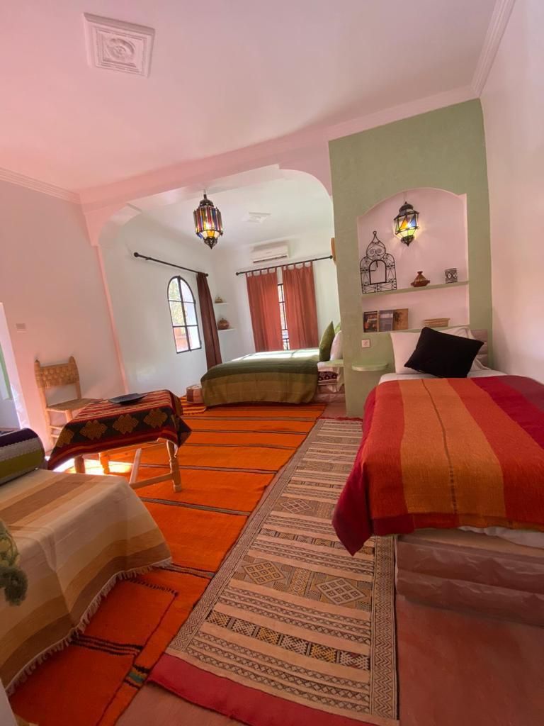 Quarto com cama, mesa e sofá. Há uma janela com cortinas e um abajur na parede.