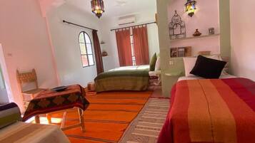 Quarto com cama, mesa e sofá. Há uma janela com cortinas e um abajur na parede.