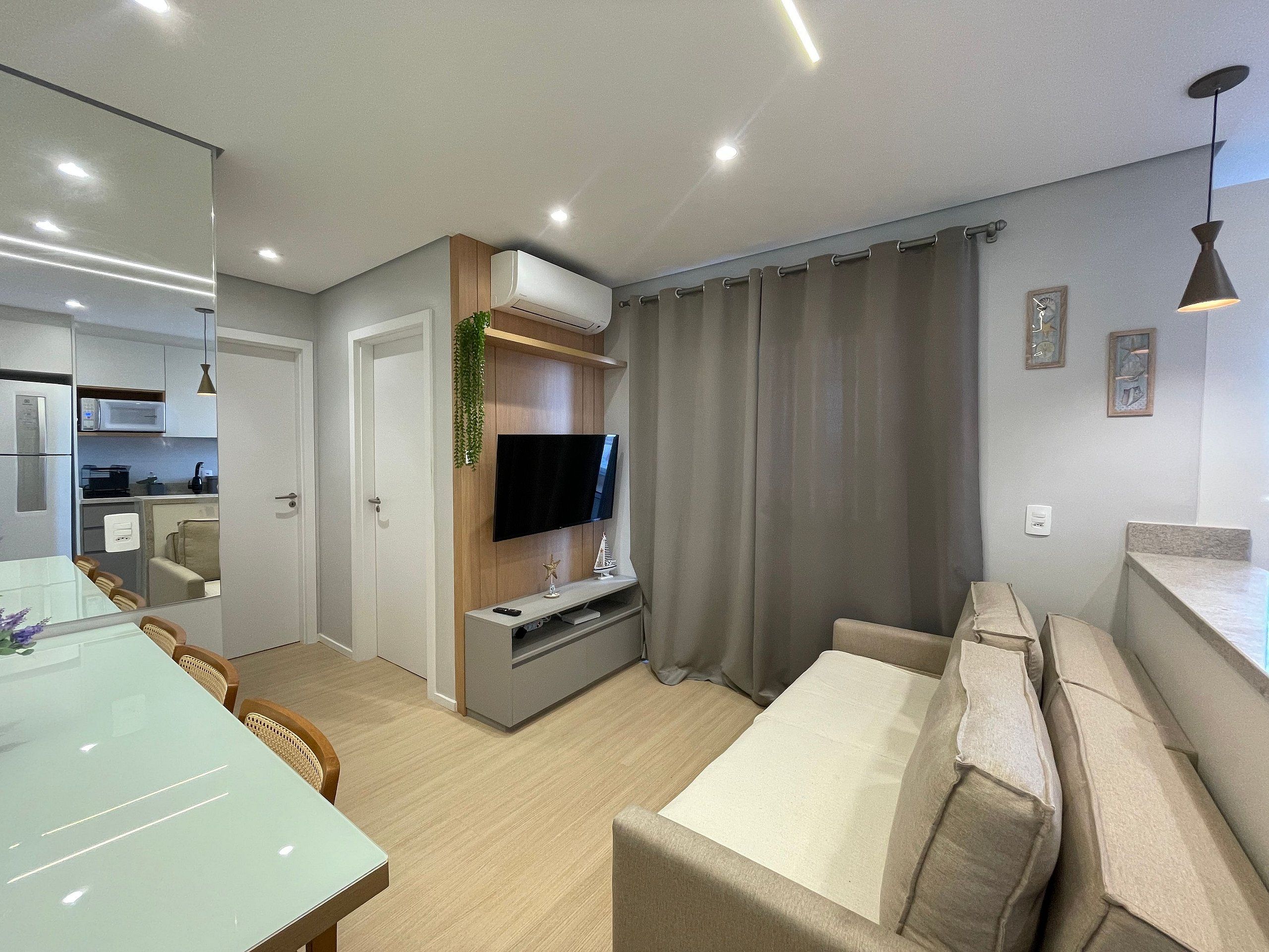 Uma sala de estar moderna com TV de tela plana, sofá, mesa de jantar com cadeiras e cozinha ao fundo.