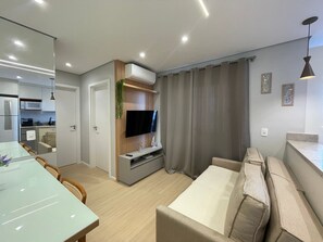 Uma sala de estar moderna com TV de tela plana, sofá, mesa de jantar com cadeiras e cozinha ao fundo.