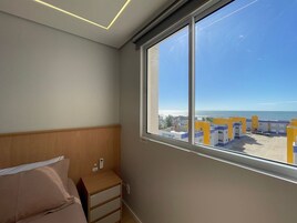 Quarto de hotel com cama, mesa de cabeceira, janela e vista para a praia e edifícios.