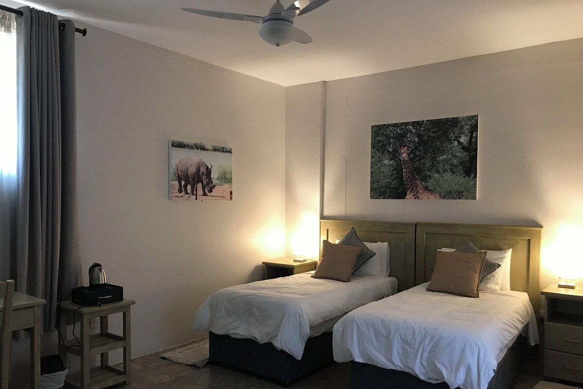 Quarto de hotel com duas camas, uma escrivaninha, uma cadeira, ventoinha no teto e obras de arte nas paredes.