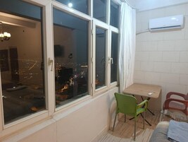 Quarto com vista para a cidade através de amplas janelas, uma pequena mesa, uma cadeira verde e uma cadeira vermelha.