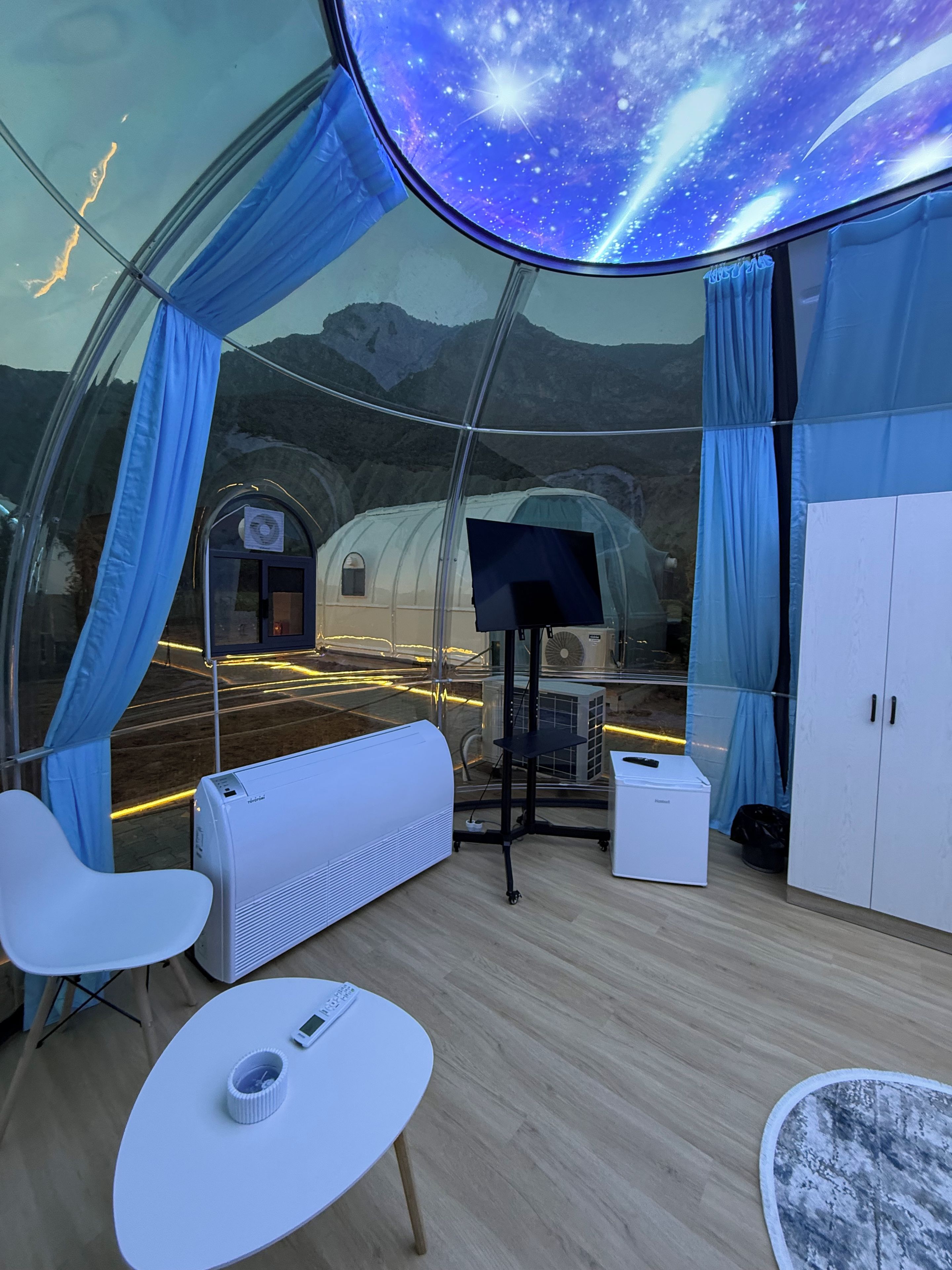 Um quarto moderno e fechado, com teto curvo exibindo um céu estrelado, área de estar com mesa e vista para uma estação de trem.