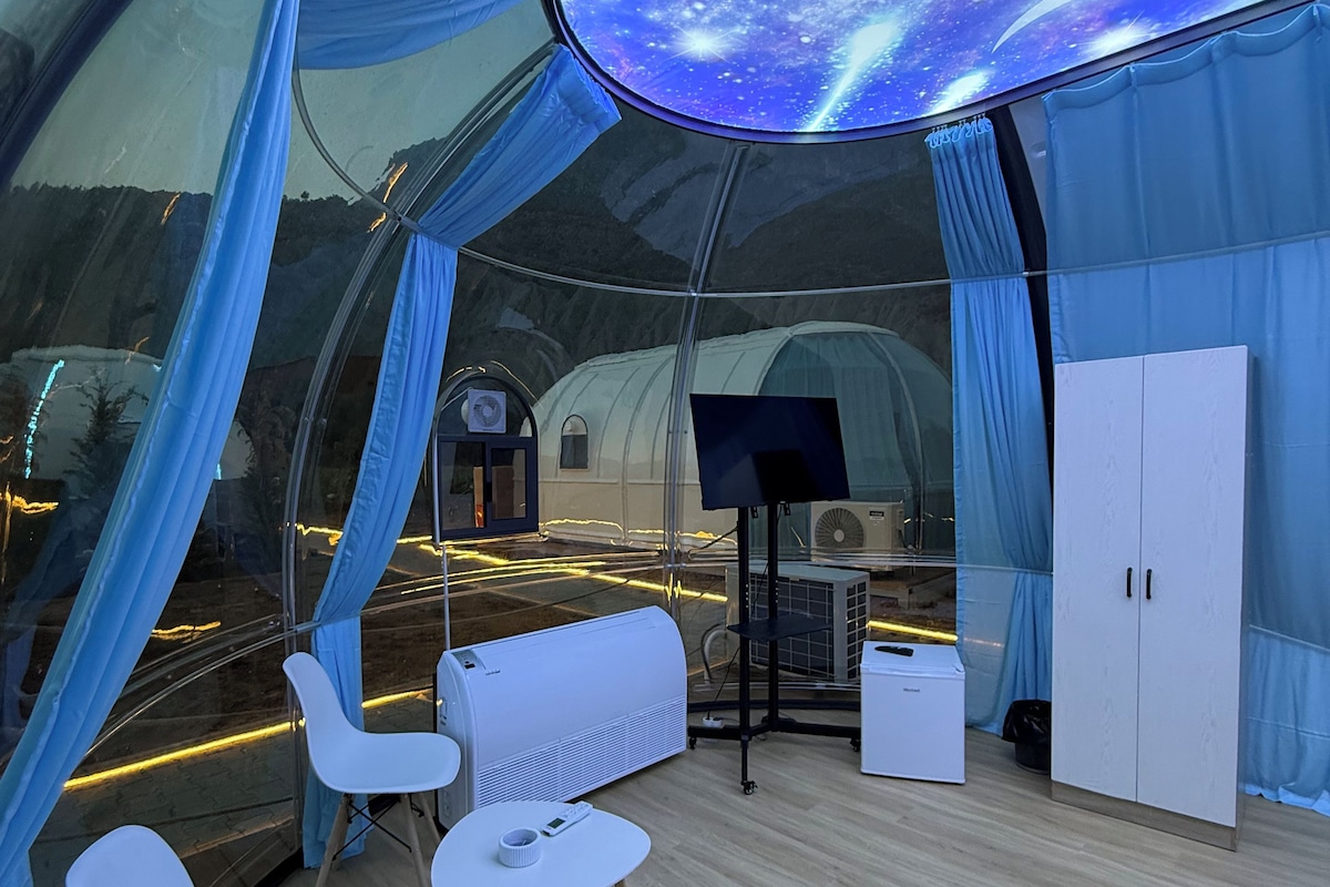 Estrutura em forma de tenda com vista para um céu estrelado através de teto transparente, mobiliário branco e uma pequena mesa com controle remoto.