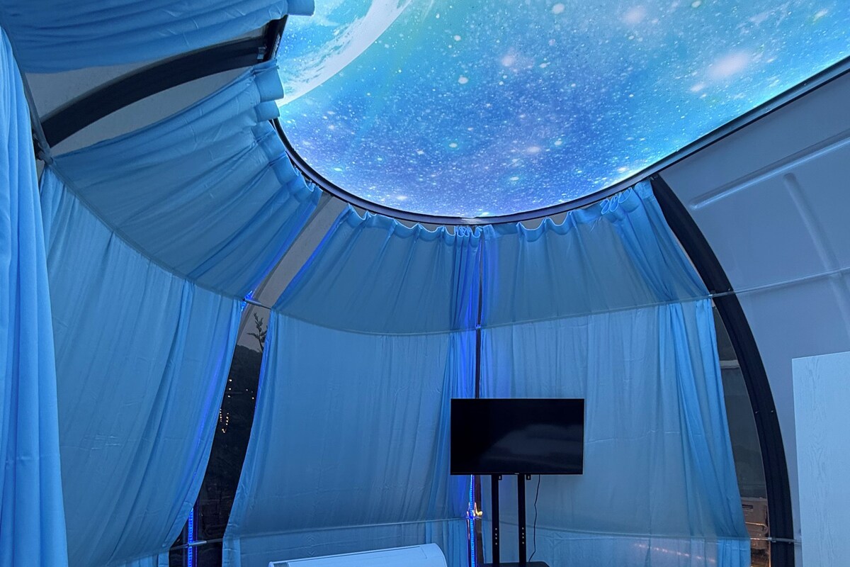 Um quarto tipo tenda com teto estrelado, uma TV de tela plana, um sofá branco, uma mesa pequena e uma cadeira branca.