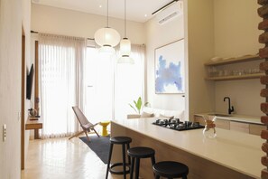 Uma cozinha moderna com uma ilha central, banquetas, um espaço de refeições com uma cadeira e uma grande janela com cortinas.