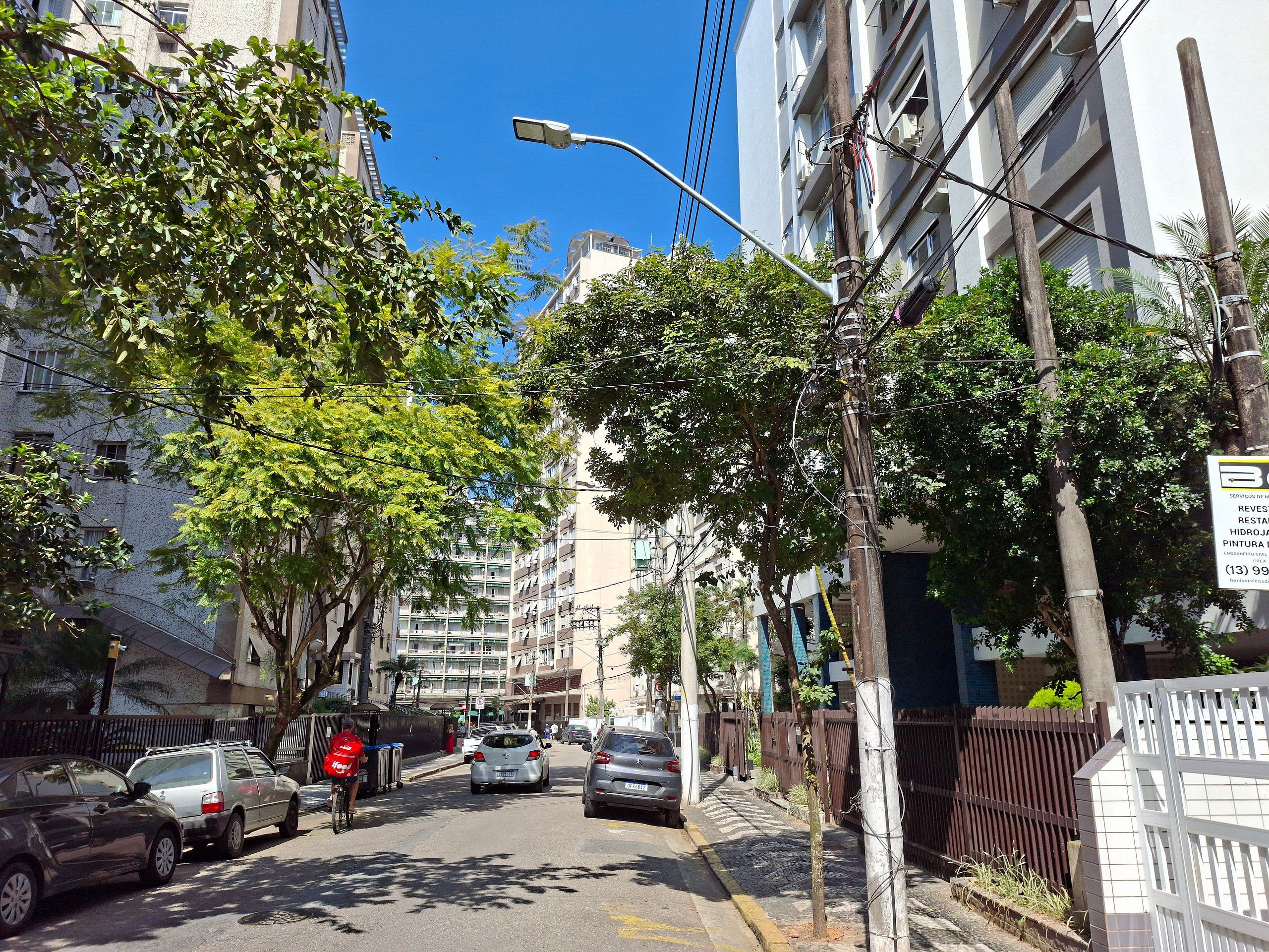 Rua urbana com carros estacionados, um ciclista e edifícios com varandas.