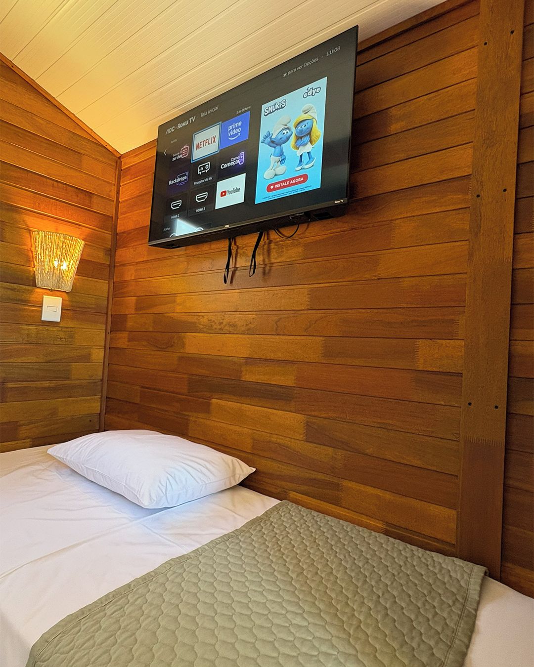 Quarto de hotel com televisão fixada na parede exibindo diversos serviços de streaming, cama com roupa de cama branca e painel de parede de madeira.