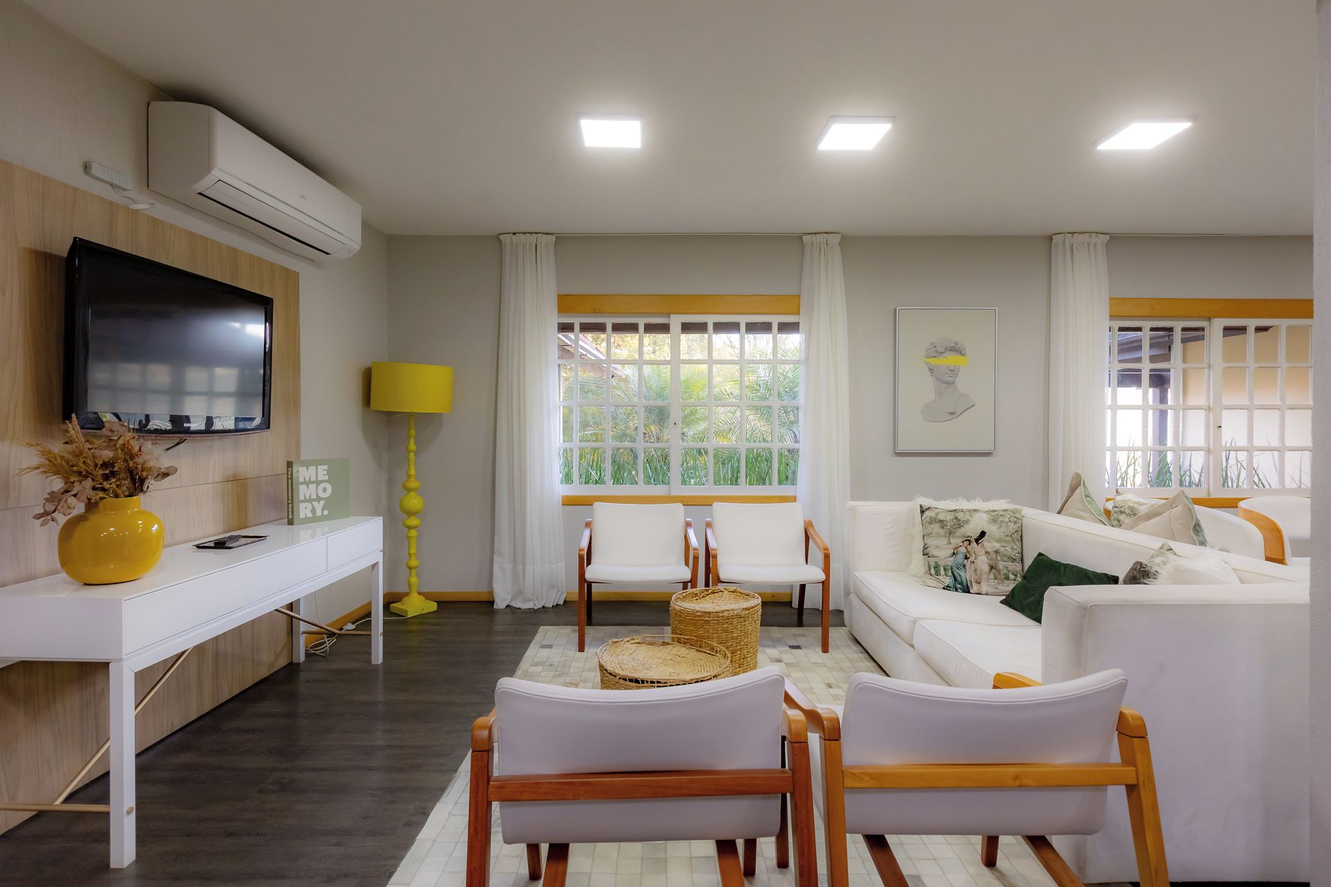 Uma sala de estar moderna com um sofá branco, duas poltronas, uma mesa de centro e uma TV fixada na parede.