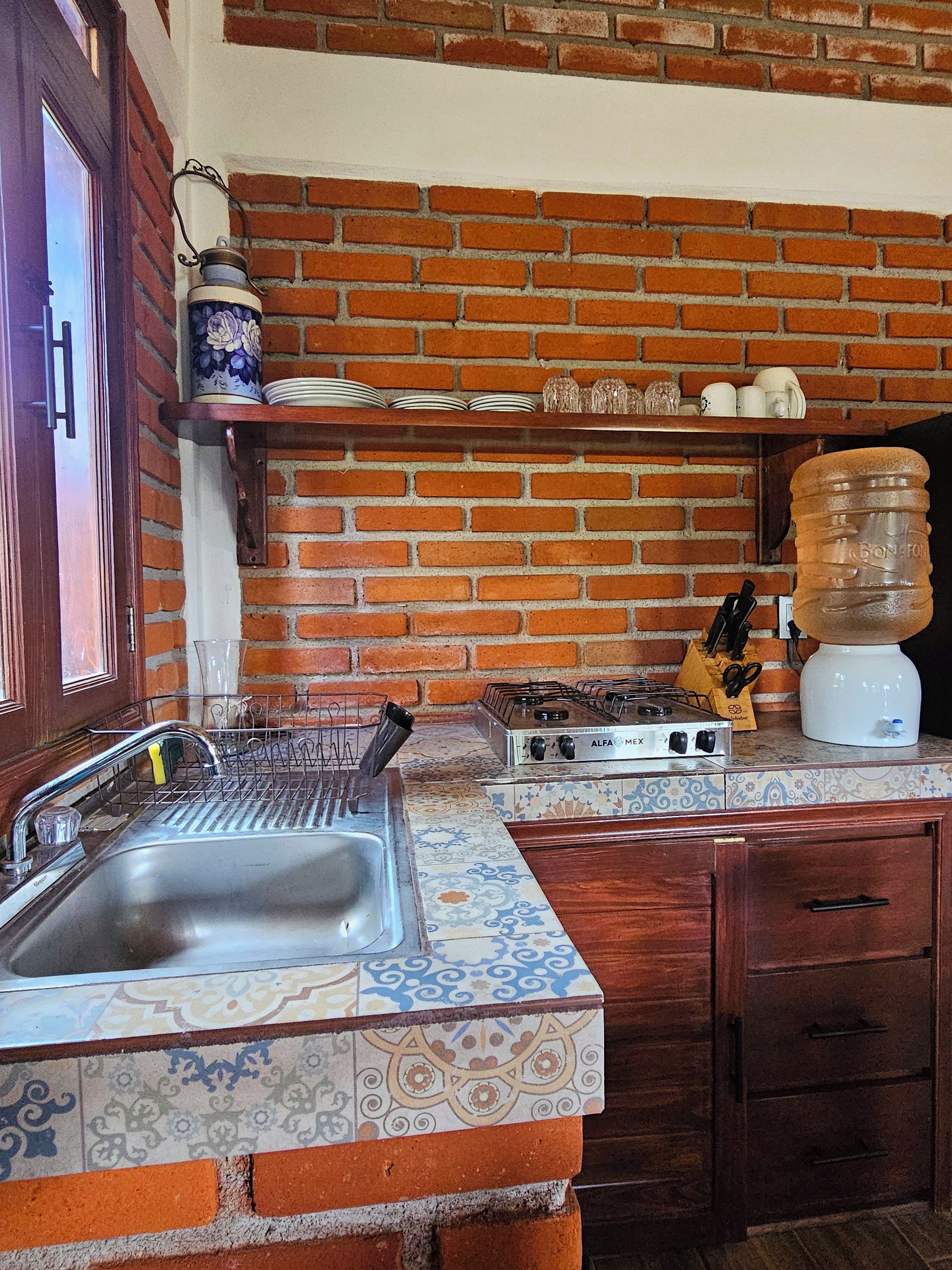 Cozinha com parede de tijolos, fogão a gás, dispenser de água e pia com escorredor de louça.