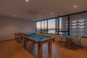 Mesa de bilhar e mesa de pebolim em um espaço com grandes janelas e vista para o horizonte da cidade.