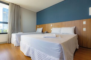 Quarto de hotel com duas camas, cabeceira de madeira, janela com cortinas e vista para edifícios.