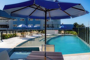 Piscina com espreguiçadeiras e guarda-sóis, cercada por um edifício com várias janelas.