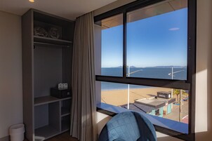 Quarto com uma grande janela com vista para a praia, uma estante e uma cadeira.