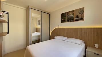 Quarto de hotel com cama, mesa de cabeceira e prateleira fixada na parede com planta em vaso.