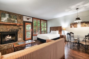Quarto aconchegante com lareira, cama, mesa de jantar com cadeiras e vista para o verde pelas janelas.