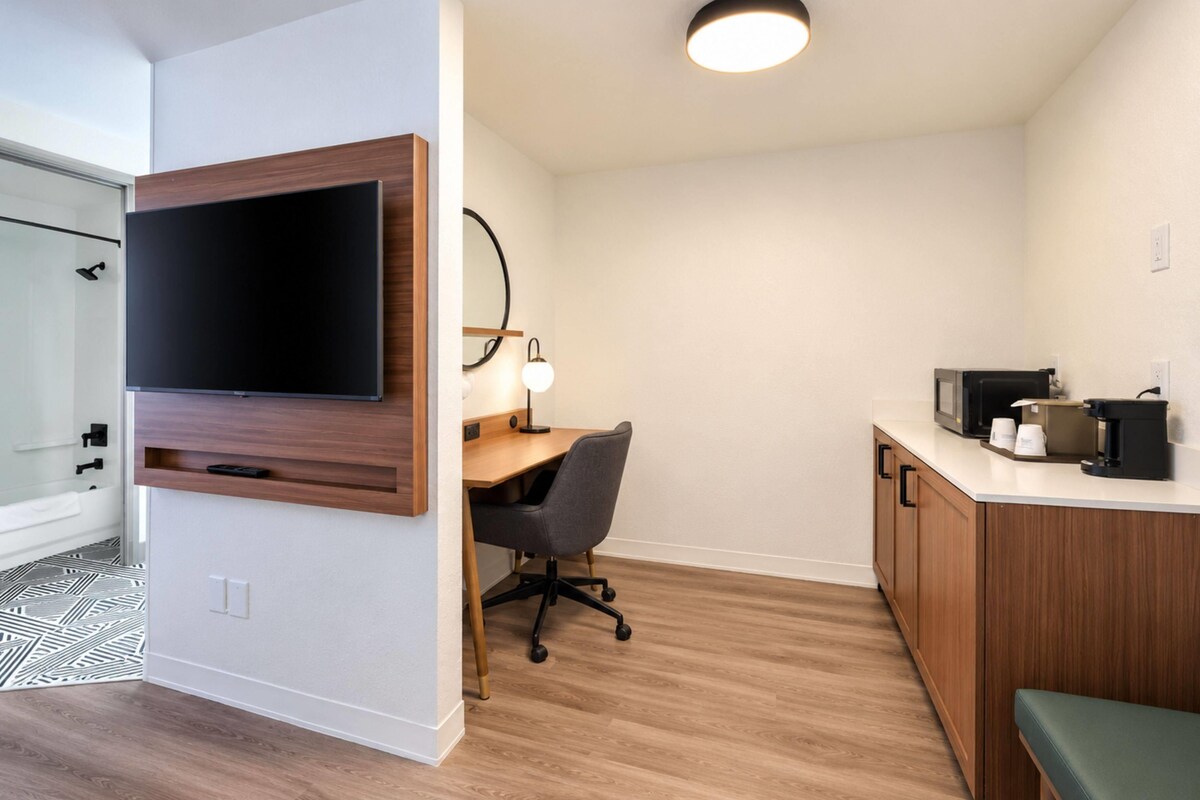 Quarto de hotel moderno com televisão de tela plana fixada em painel de madeira, uma escrivaninha com cadeira, uma pequena mesa com luminária e uma cozinha compacta com micro-ondas e cafeteira.