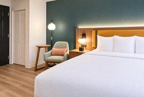 Quarto de hotel com cama, poltrona, mesa de cabeceira, luminária e guarda-roupa.