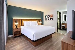 Quarto de hotel com cama, mesas de cabeceira, televisão fixada na parede e porta para o banheiro.