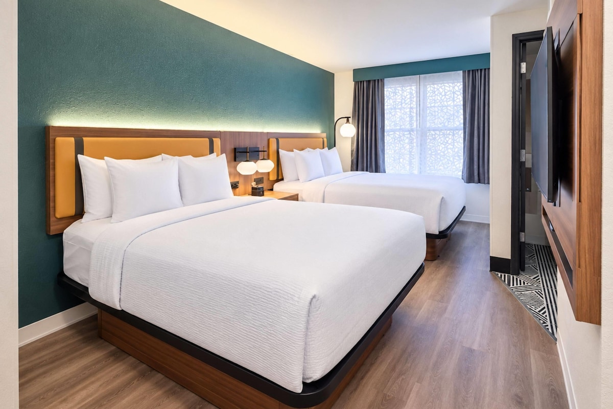 Quarto de hotel com duas camas, cabeceira de madeira, mesa de cabeceira com abajur e janela com cortinas.