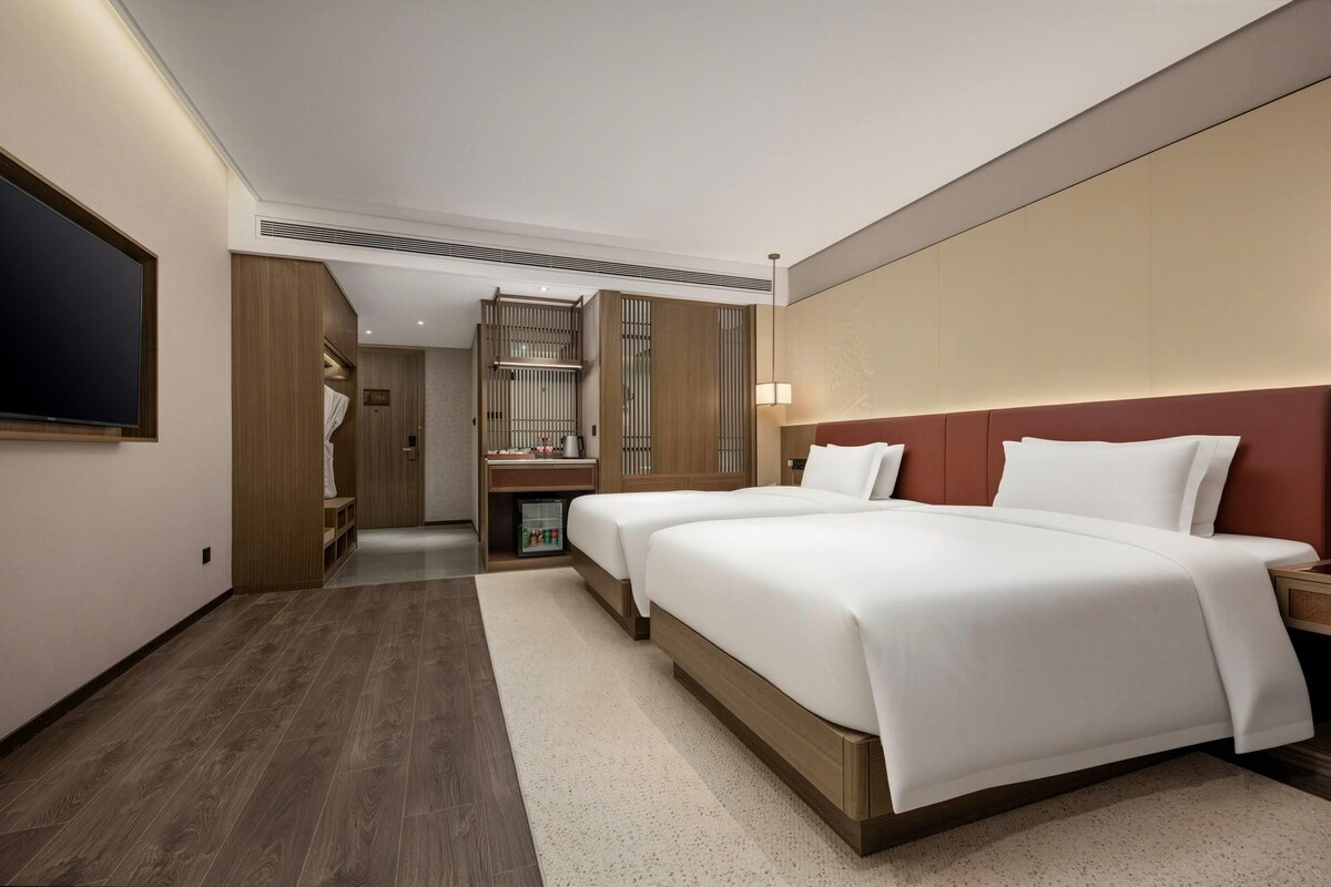 Quarto de hotel com duas camas, televisão, minibar e banheiro com chuveiro.