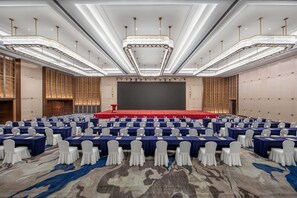 Uma sala de conferências com mesas e cadeiras dispostas em fileiras, um palco e uma tela grande.