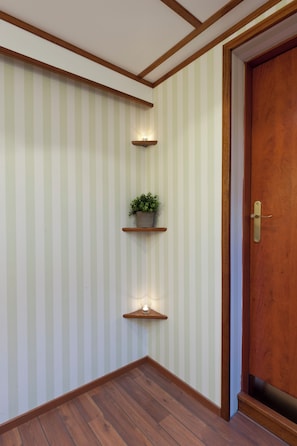 Canto de um quarto com papel de parede listrado, uma porta de madeira e duas prateleiras com uma planta e uma vela.