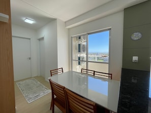 Uma sala de jantar com mesa e cadeiras, janela com aparelhos de ar condicionado e vista para edifícios.