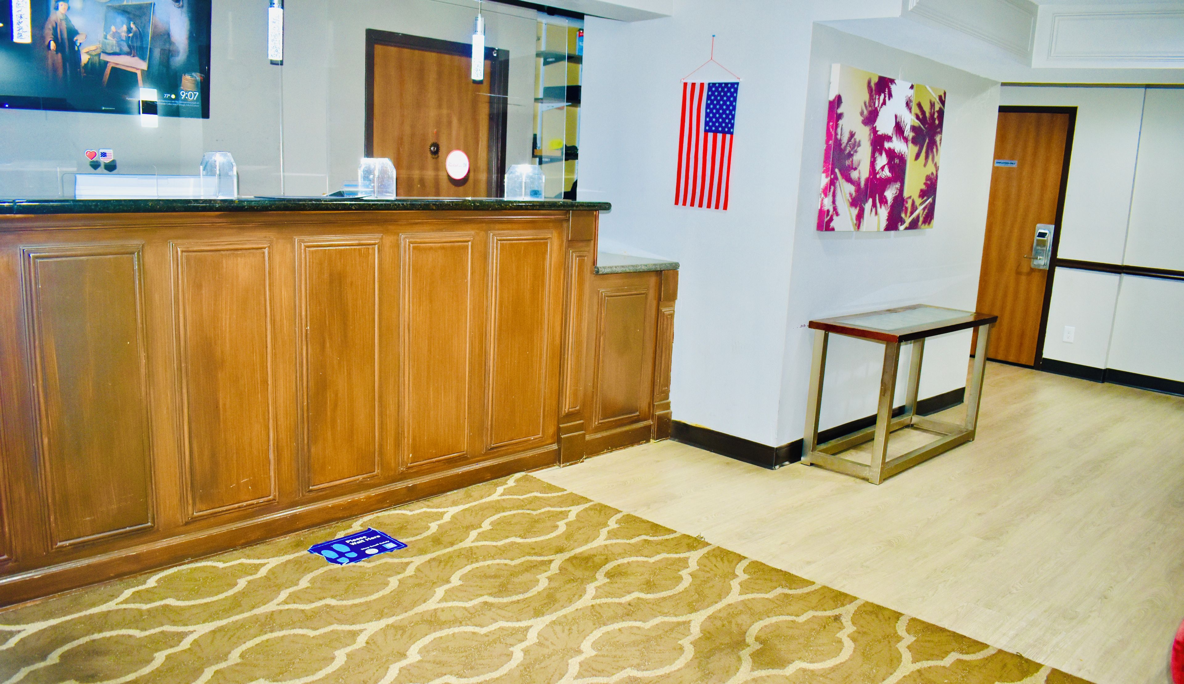 Um saguão de hotel com um balcão de recepção de madeira, uma pequena mesa, uma bandeira americana e um quadro na parede.
