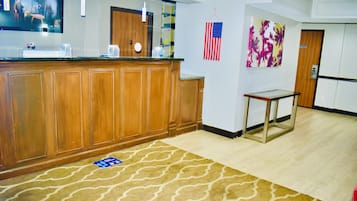 Um saguão de hotel com um balcão de recepção de madeira, uma pequena mesa, uma bandeira americana e um quadro na parede.