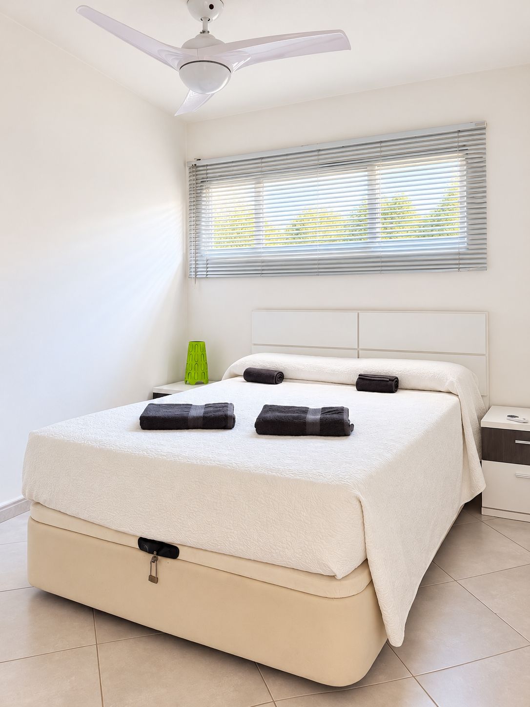 Cama bem arrumada com cabeceira branca, ventilador de teto e janela com persianas.