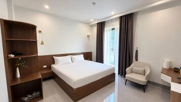Quarto de hotel moderno com uma cama grande, uma mesinha de cabeceira de madeira, uma cadeira, uma escrivaninha e uma janela com cortinas.