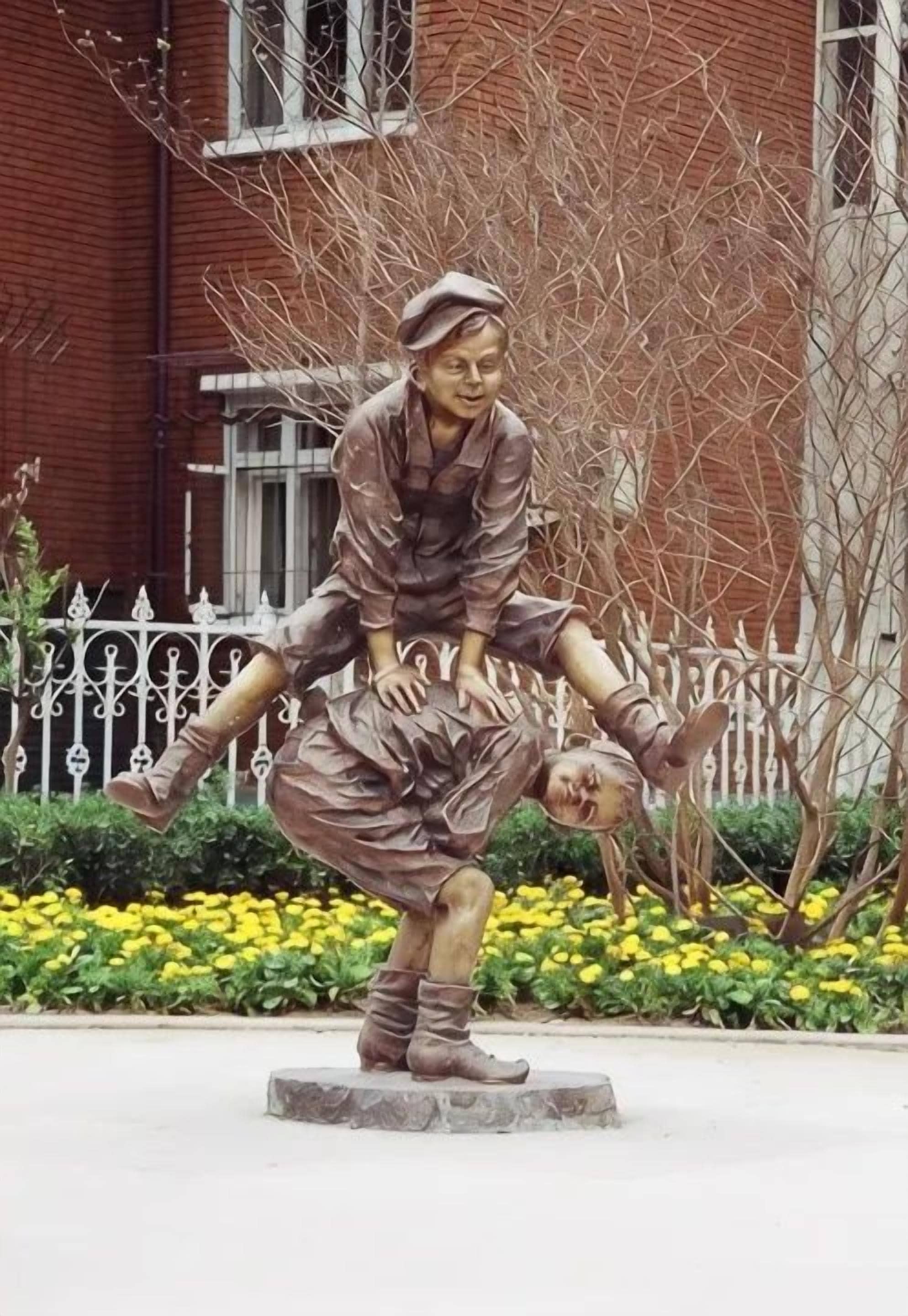Estátua de bronze de uma pessoa em pose dinâmica, com um edifício de tijolos e uma grade de metal com flores amarelas ao fundo.
