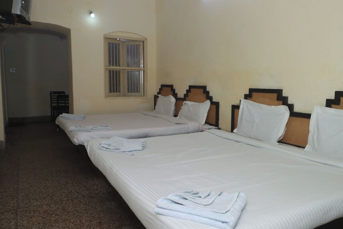 Quarto de hotel com três camas, uma janela com grades, uma mesinha e uma cadeira.