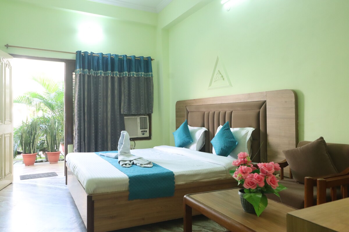 Quarto de hotel com cama, mesinha, cadeira, planta em vaso e janela com cortinas.
