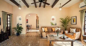 Uma sala de estar ampla, com teto alto, ventilador de teto, um sofá grande, mesa de centro e um espaço de refeições ao fundo.