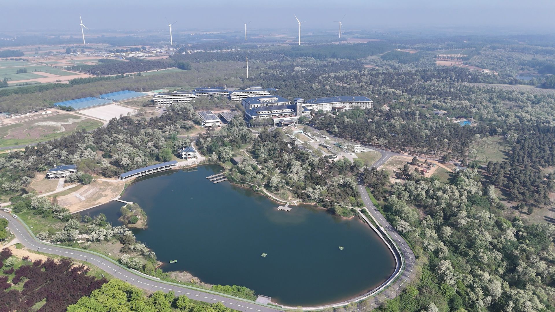 Vista aérea de um campus com um lago, edifícios e turbinas eólicas ao fundo.