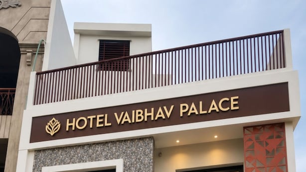 Um hotel de dois andares com fachada moderna, uma placa de destaque com a inscrição 'HOTEL VAIBHAV PALACE' e entrada de vidro que dá acesso a uma área de recepção.