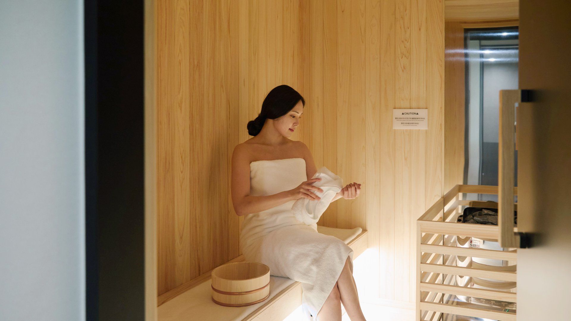 Uma pessoa em uma sauna de madeira com toalha, um banco de madeira e um aviso.