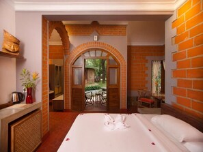 Quarto de hotel com cama, escrivaninha, cadeira e vista para um jardim através de uma porta arqueada.