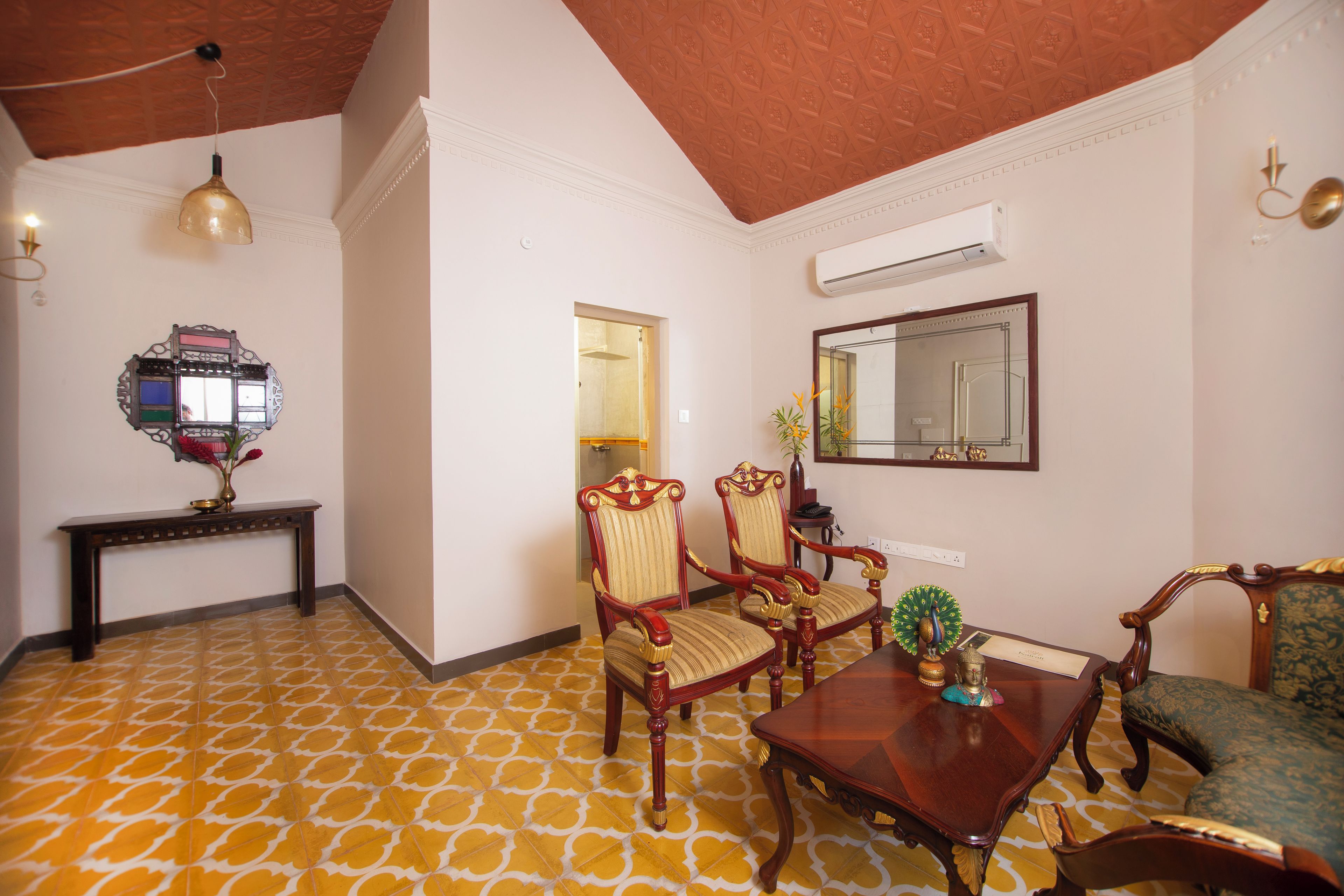 Quarto com ladrilhos amarelados e brancos, mesa de centro de madeira, duas cadeiras ornamentadas, espelho e um vaso decorativo.