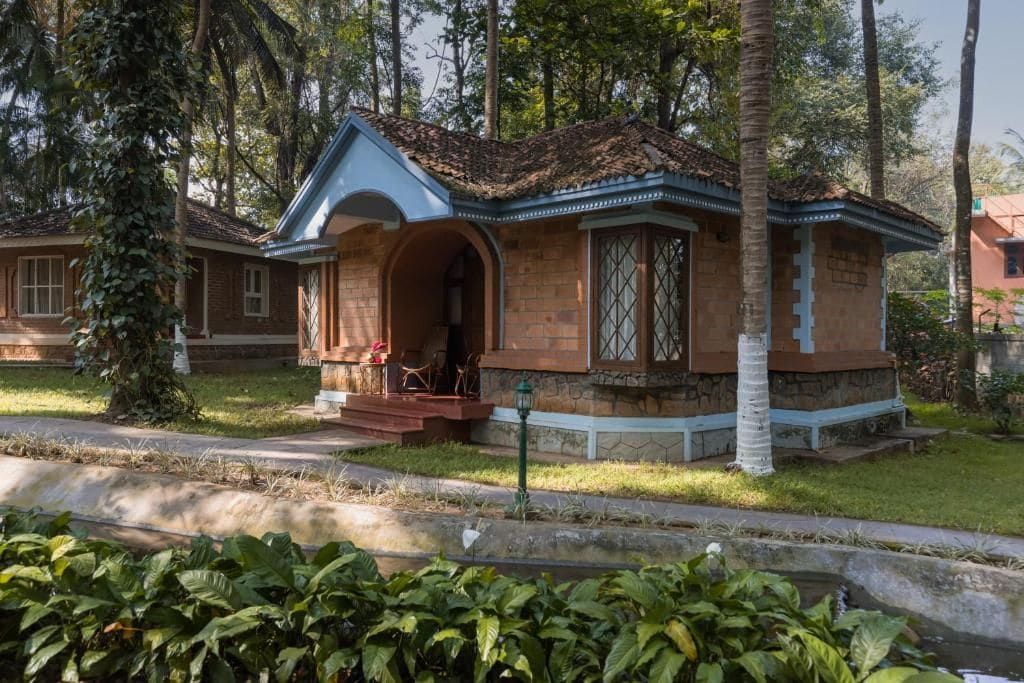 Uma casa pequena e térrea, com telhado de telhas, varanda com cadeiras e alicerce de pedra.